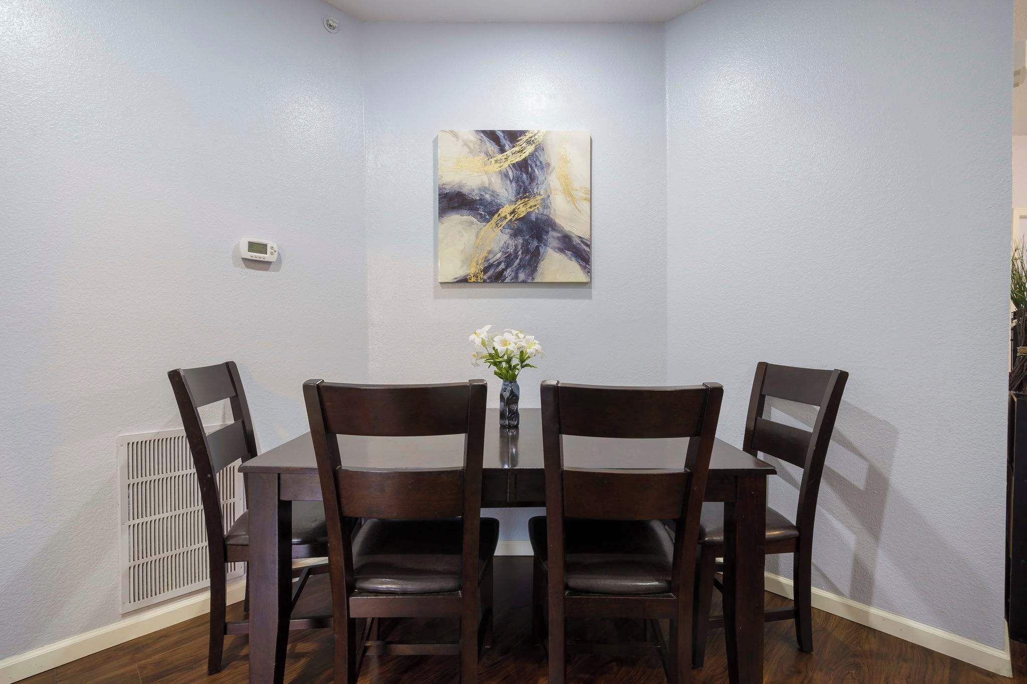 Uma área de refeições com uma mesa e quatro cadeiras, um termostato fixado na parede e uma pintura sobre a mesa.