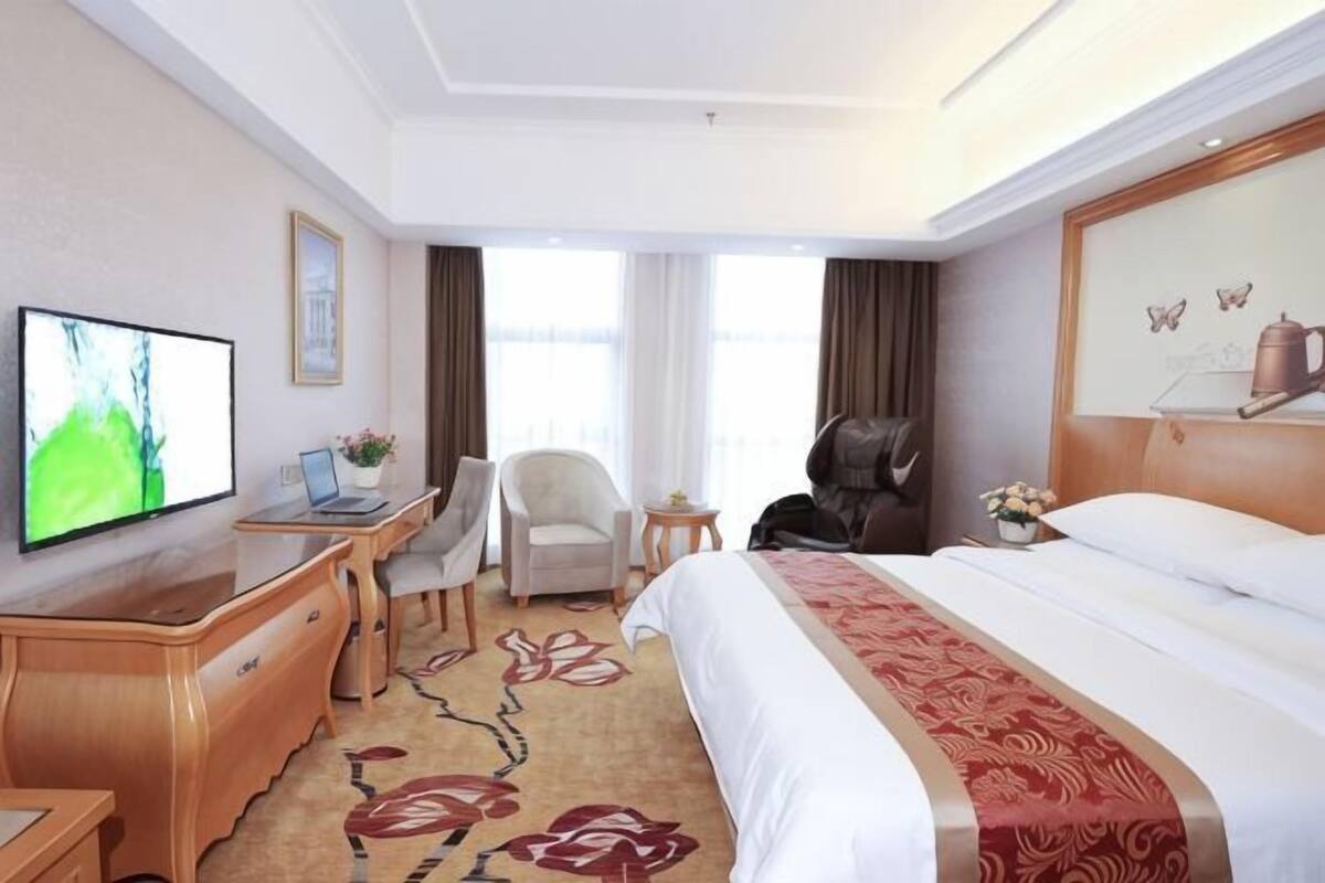 Quarto de hotel com cama, escrivaninha, cadeira, televisão e janela com cortinas.