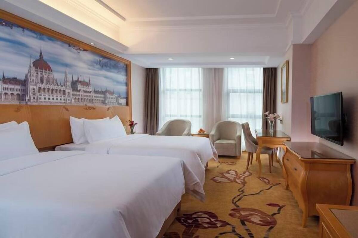 Quarto de hotel com duas camas, uma escrivaninha, uma cadeira, uma televisão e um quadro de um edifício.