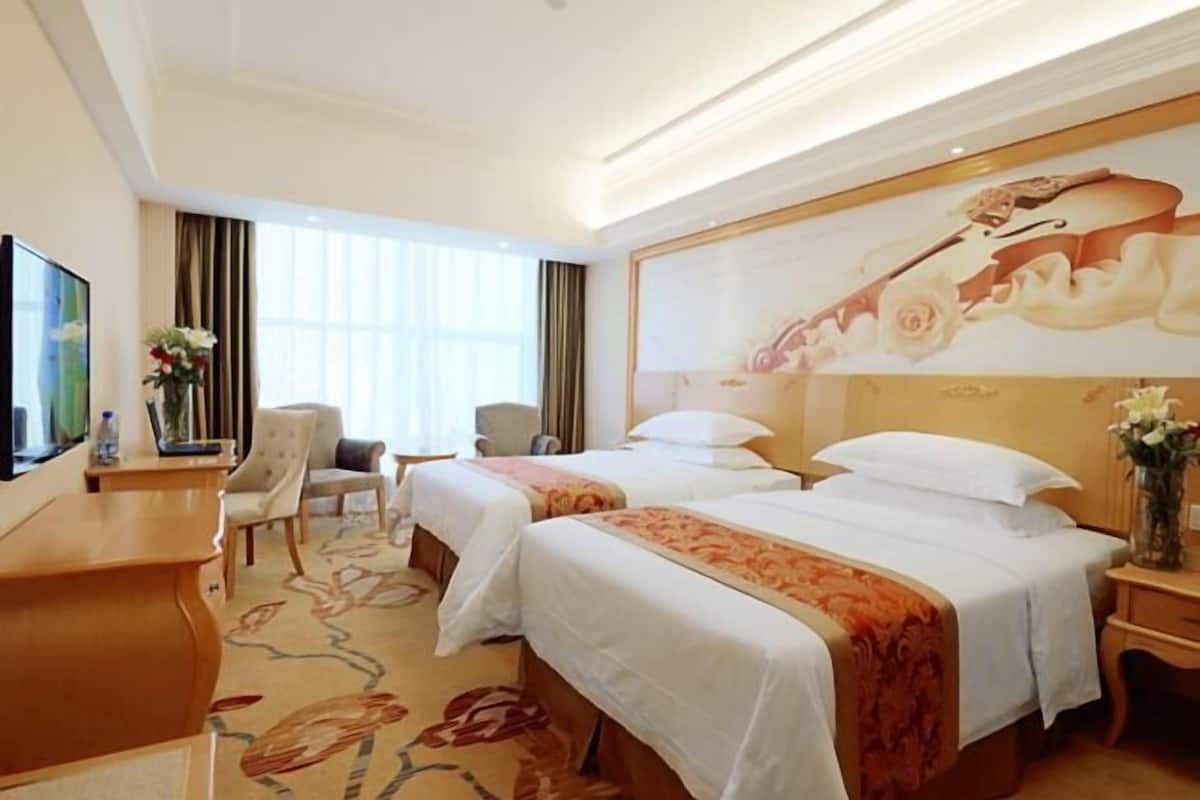 Quarto de hotel com duas camas, uma escrivaninha, uma cadeira, uma televisão e um quadro grande na parede.