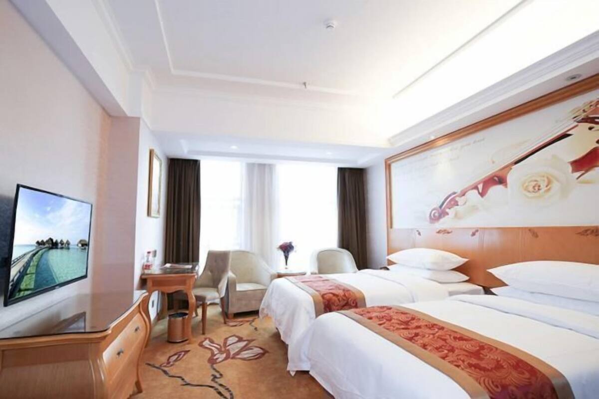 Quarto de hotel com televisão, duas camas, uma secretária, uma cadeira e uma janela grande.