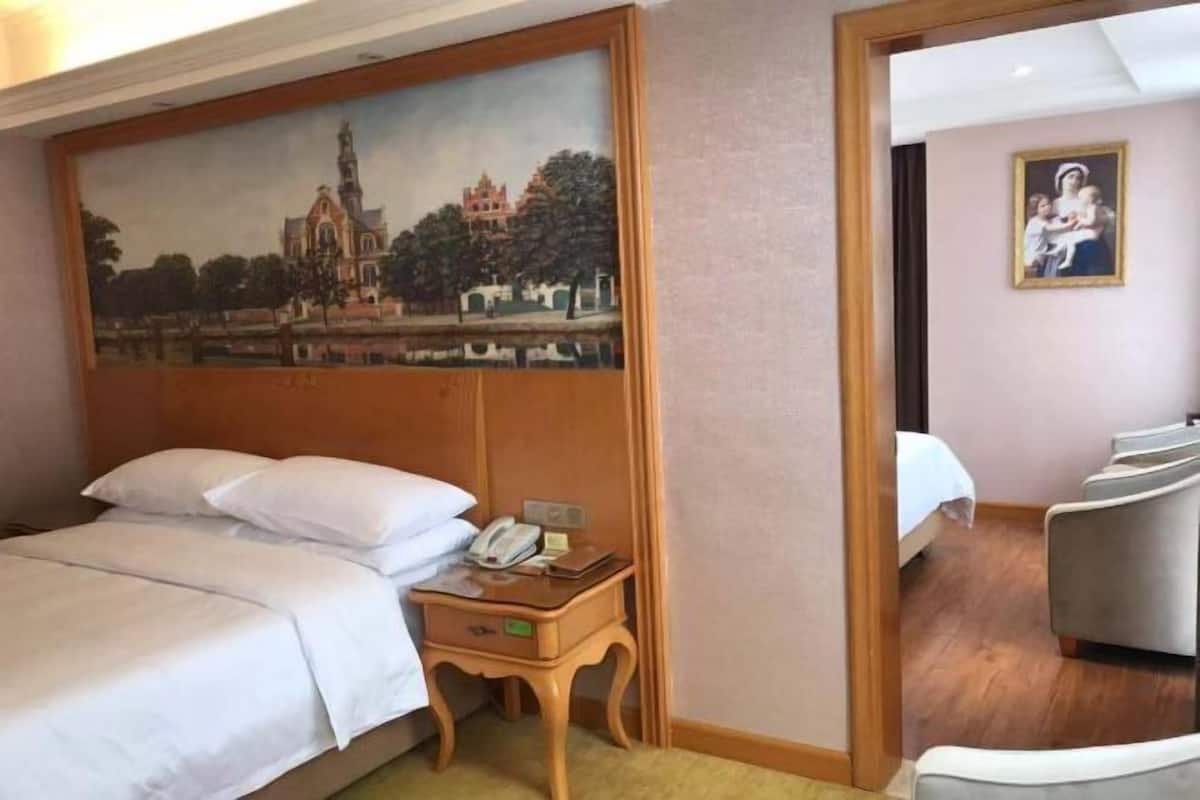Quarto de hotel com cama, mesa de cabeceira com telefone, quadro de um edifício e uma cadeira.