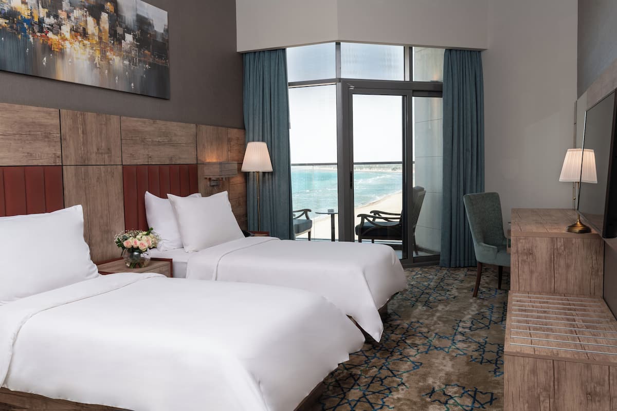 Quarto de hotel com duas camas, uma escrivaninha, uma cadeira, uma janela ampla com vista para o oceano e um quadro na parede.