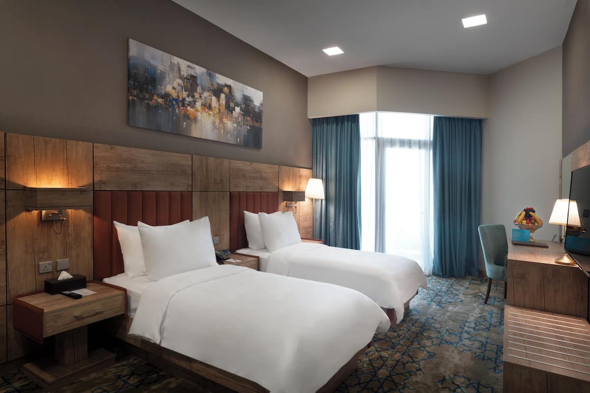 Quarto de hotel com duas camas, cabeceira de madeira, uma grande pintura abstrata, escrivaninha com luminária e vista da cidade através de amplas janelas.