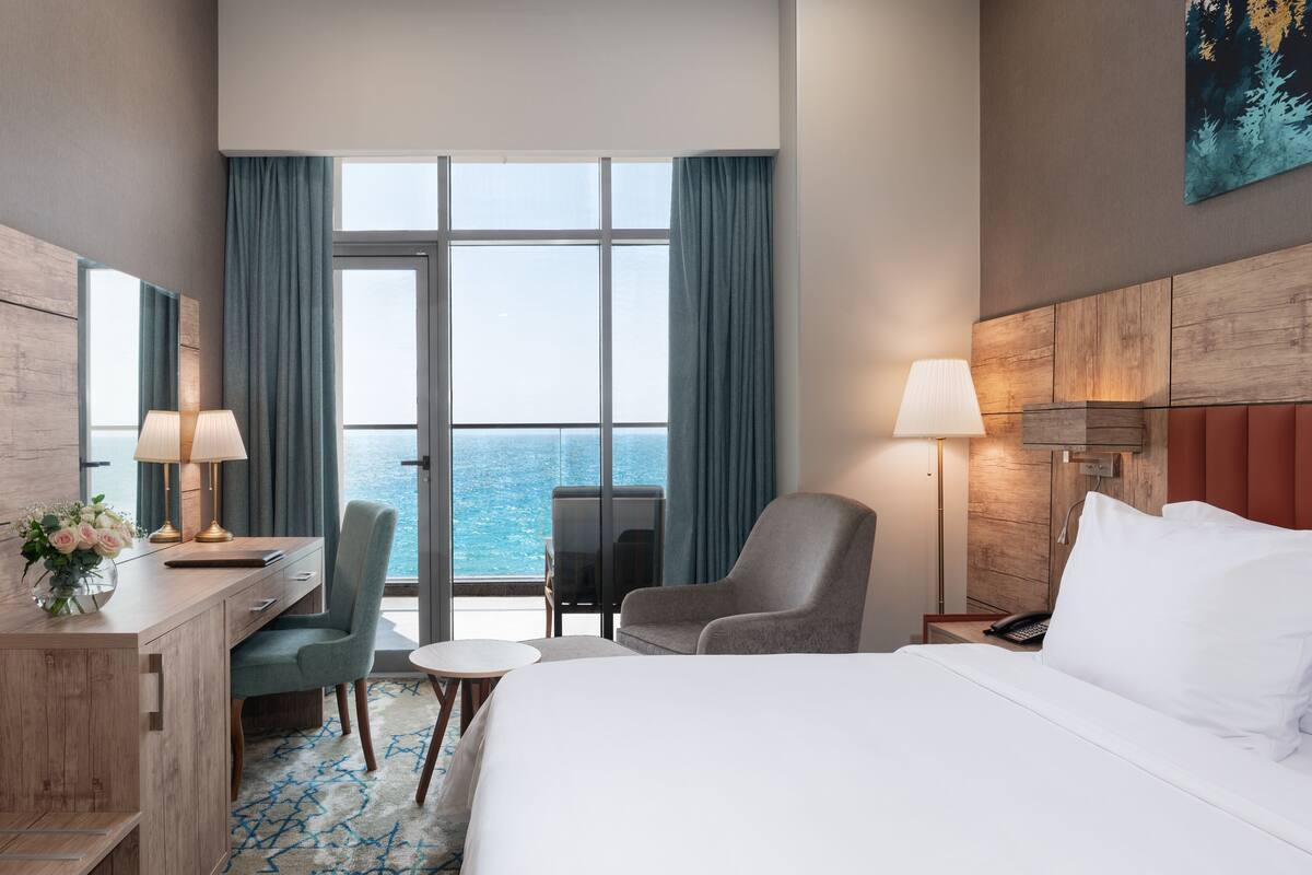 Quarto de hotel com uma cama grande, uma escrivaninha com computador, uma cadeira, uma mesa pequena e uma televisão. Há uma janela com vista para o oceano.