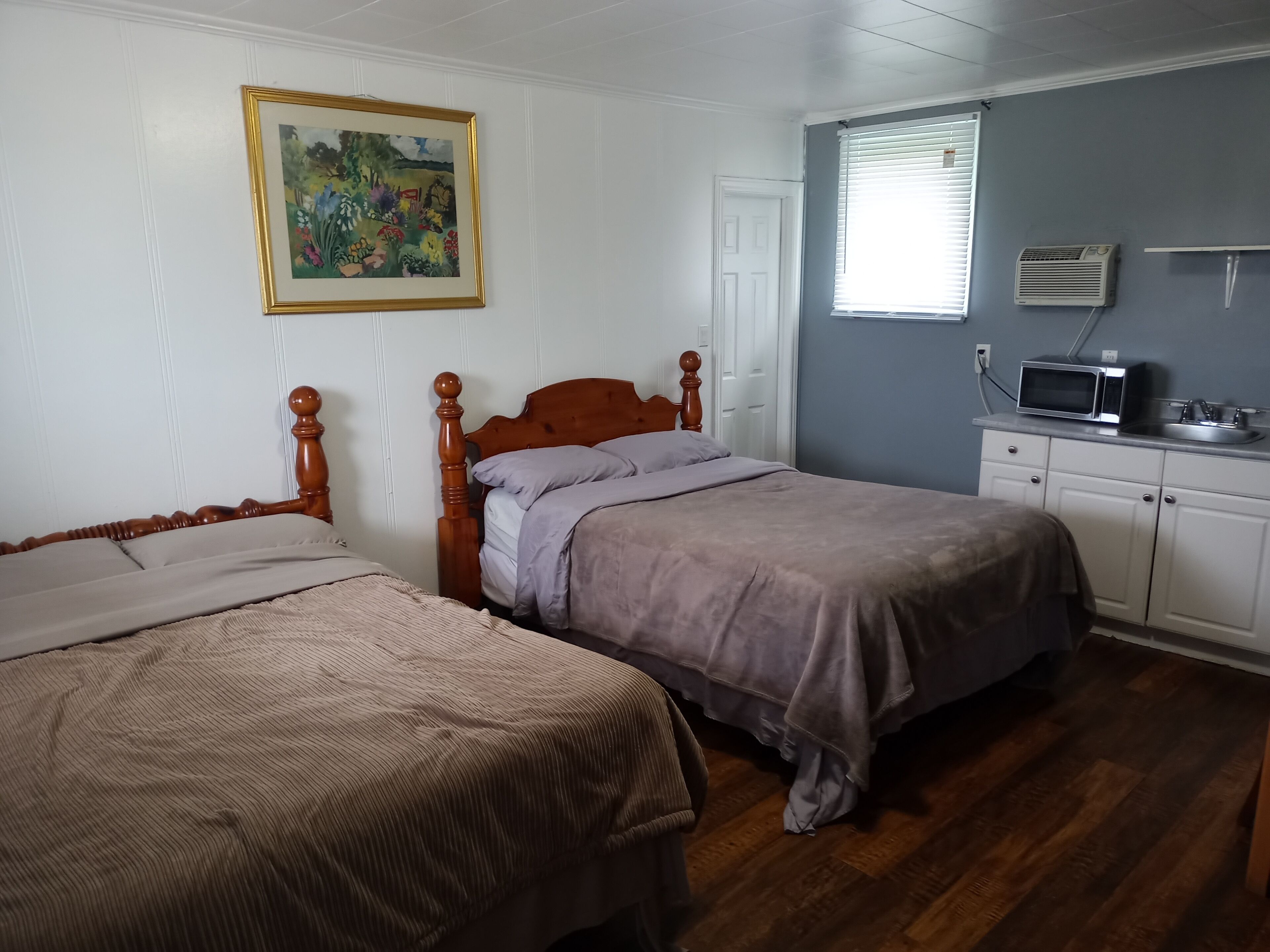 Quarto com duas camas, um quadro na parede, janela com persianas e uma cozinha compacta com micro-ondas.