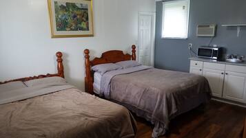 Quarto com duas camas, um quadro na parede, janela com persianas e uma cozinha compacta com micro-ondas.