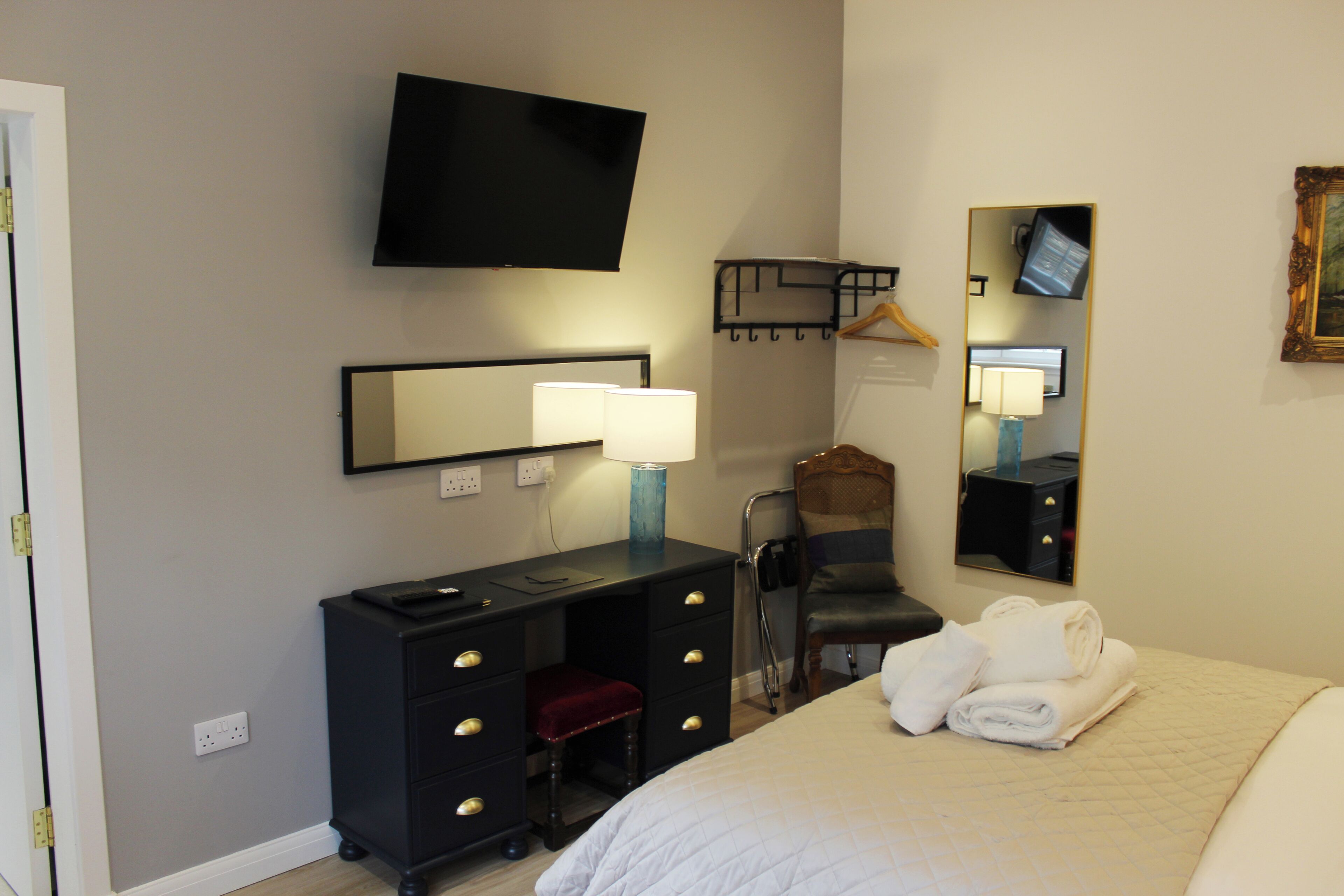 Quarto de hotel com uma cama, uma escrivaninha com cadeira, uma televisão na parede, um espelho e uma prateleira na parede com uma TV.