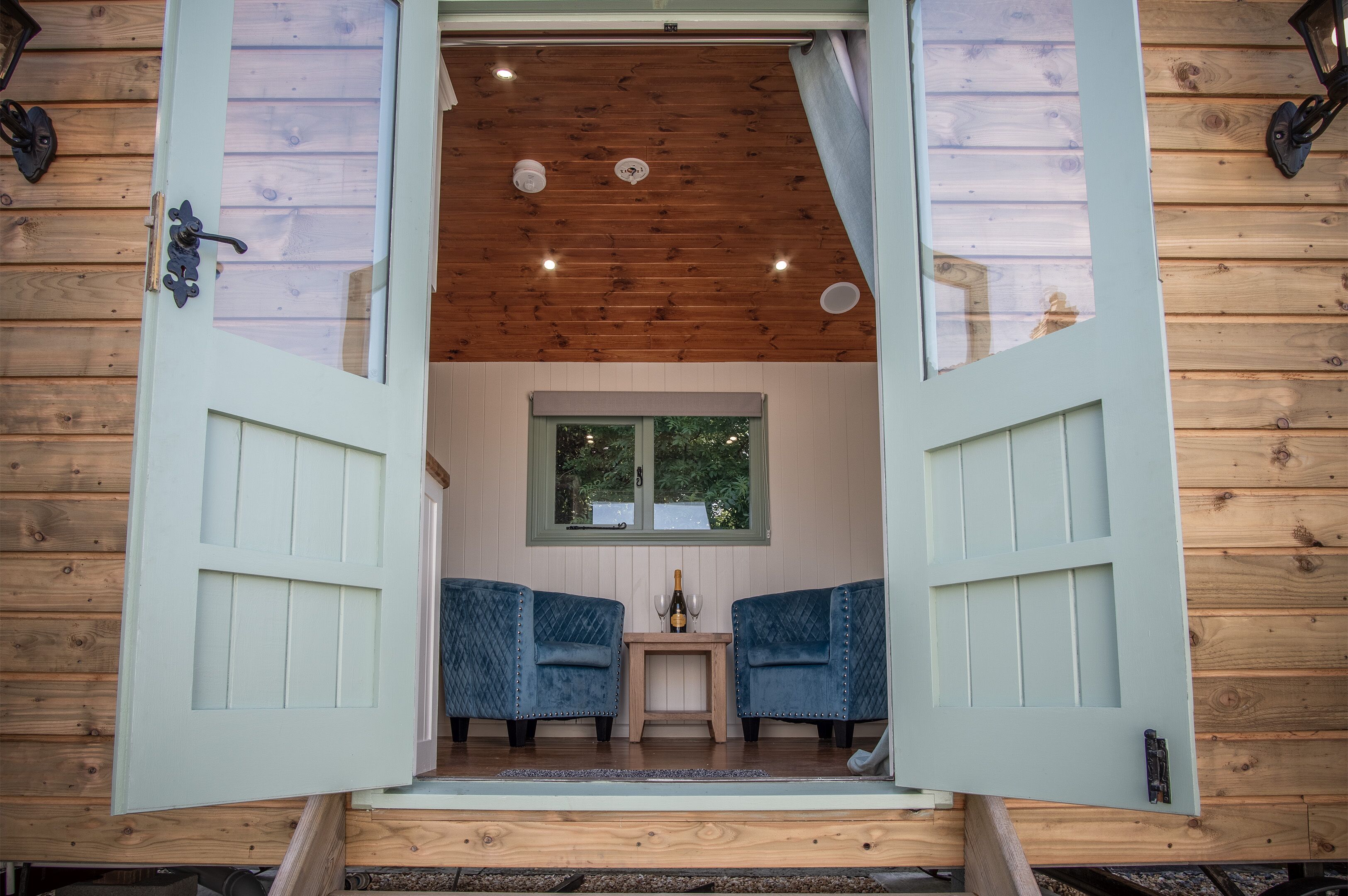 Entrada de uma cabana de madeira com a porta aberta, mostrando uma pequena área de estar com duas cadeiras, uma mesa, uma janela e uma garrafa de vinho.