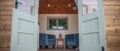 Entrada de uma cabana de madeira com a porta aberta, mostrando uma pequena área de estar com duas cadeiras, uma mesa, uma janela e uma garrafa de vinho.