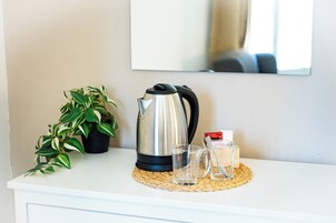 Uma kitchenette com uma chaleira de aço inoxidável, dois copos, um pequeno recipiente e uma planta em vaso sobre um balcão branco.