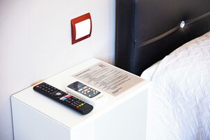 Cama com edredom branco e controle remoto sobre uma mesinha de cabeceira branca.