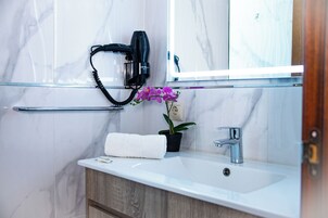 Casa de banho com parede de mármore, lavatório branco, toalha, orquídea num vaso e secador de cabelo fixado na parede.