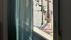 Visão através de uma janela com cortinas leves, mostrando uma rua com palmeiras, carros estacionados e céu claro.