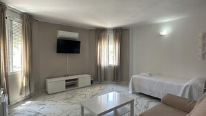 Quarto de hotel moderno com piso de mármore, TV de tela plana na parede, cama com edredom branco, sofá bege e uma pequena mesa branca.
