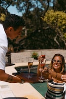 Cenário à beira da piscina com uma mesa de madeira e duas pessoas brindando com bebidas.