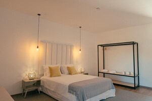 Quarto com cama, luminárias de cabeceira, mesa de cabeceira, um espelho grande e prateleira com toalhas.