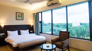 Quarto de hotel com cama, cadeira, mesa pequena, ventoinha no teto, ar condicionado e uma grande janela com vista para áreas verdes e edifícios.