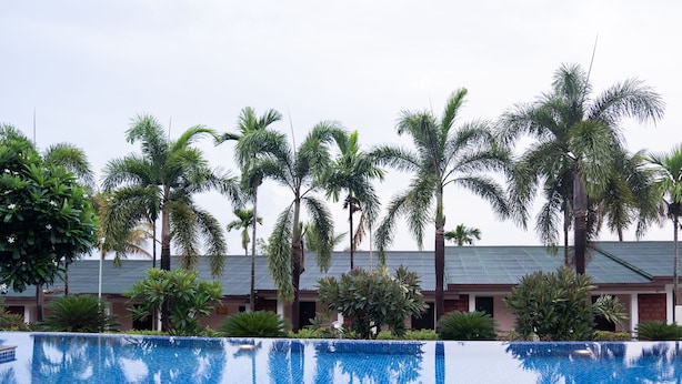 Piscina com borda azulejada, rodeada por palmeiras e um edifício com telhado de metal corrugado.