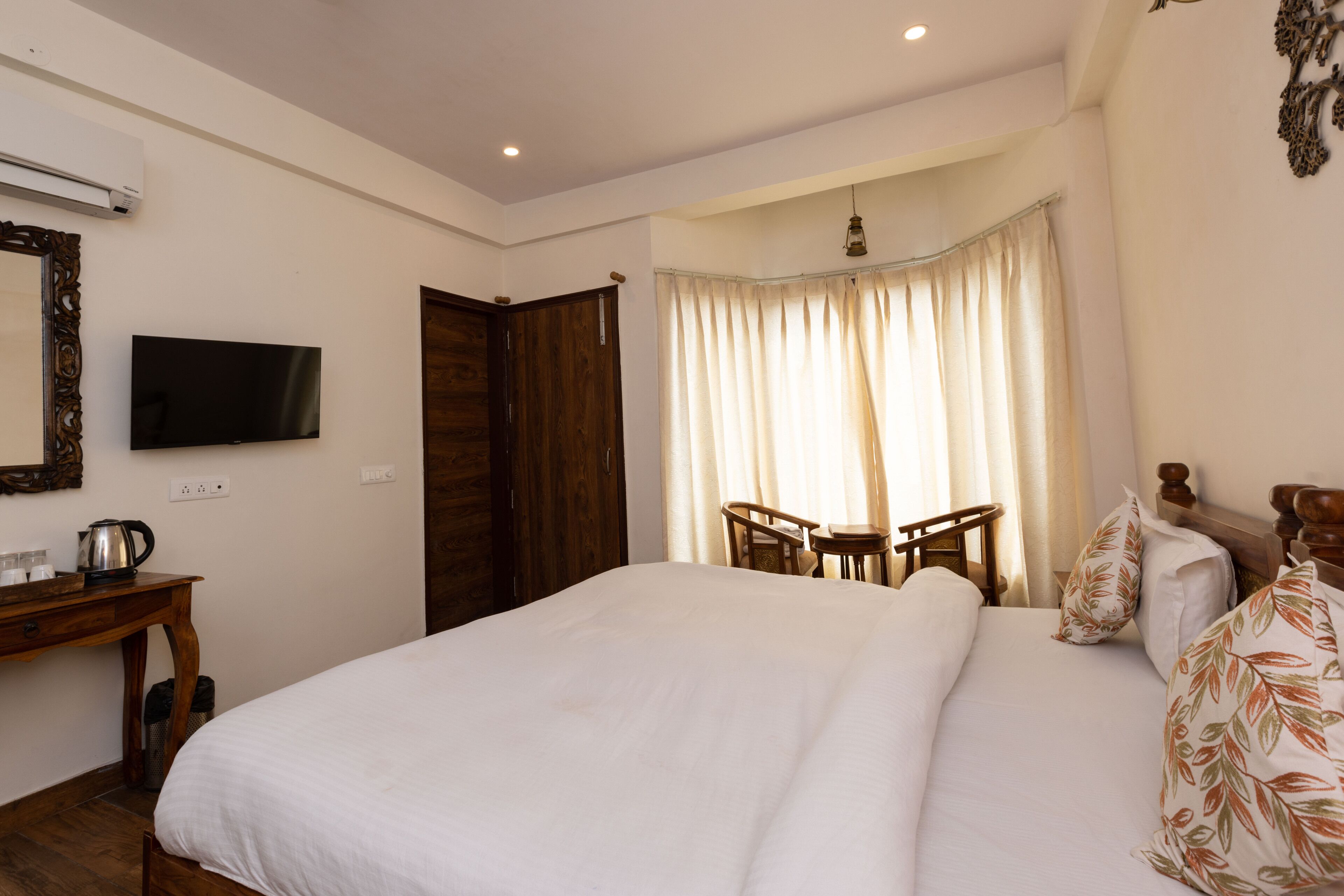 Quarto com uma cama grande, televisão na parede, escrivaninha de madeira com chaleira e janela com cortinas.