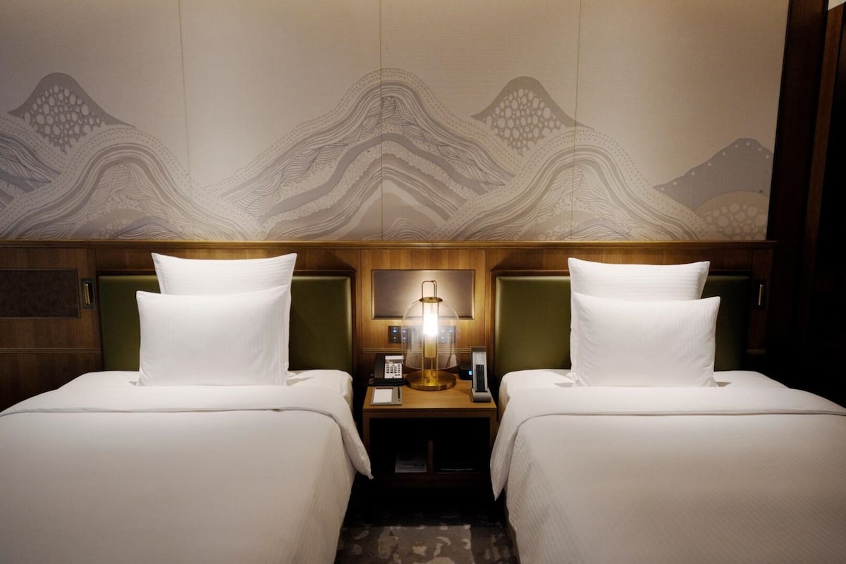 Quarto de hotel com duas camas, uma mesa de cabeceira com abajur e uma parede com padrão de ondas.
