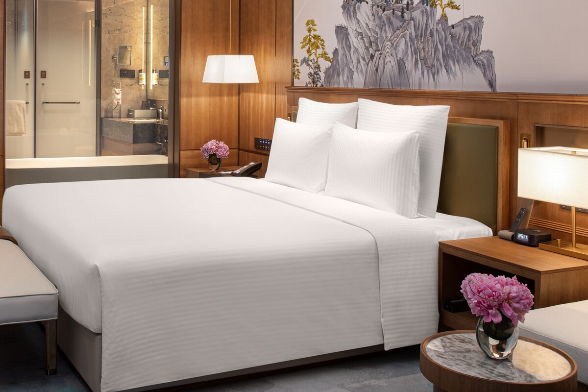 Quarto de hotel com uma cama grande, mesa de cabeceira com abajur, criado-mudo com telefone e banheiro visível através de uma porta de vidro.