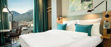 Quarto de hotel com uma cama grande, uma escrivaninha e uma cadeira. Vê-se montanhas e uma paisagem urbana pela janela.