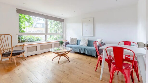 Um quarto claro com piso de madeira, uma mesa de vidro, um sofá e cadeiras vermelhas.