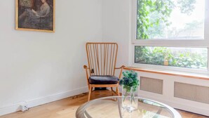 Quarto com cadeira de madeira, mesa de vidro, quadro na parede e janela com vista para árvores.