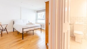 Quarto com cama, piso de madeira, janela e banheiro com vaso sanitário.