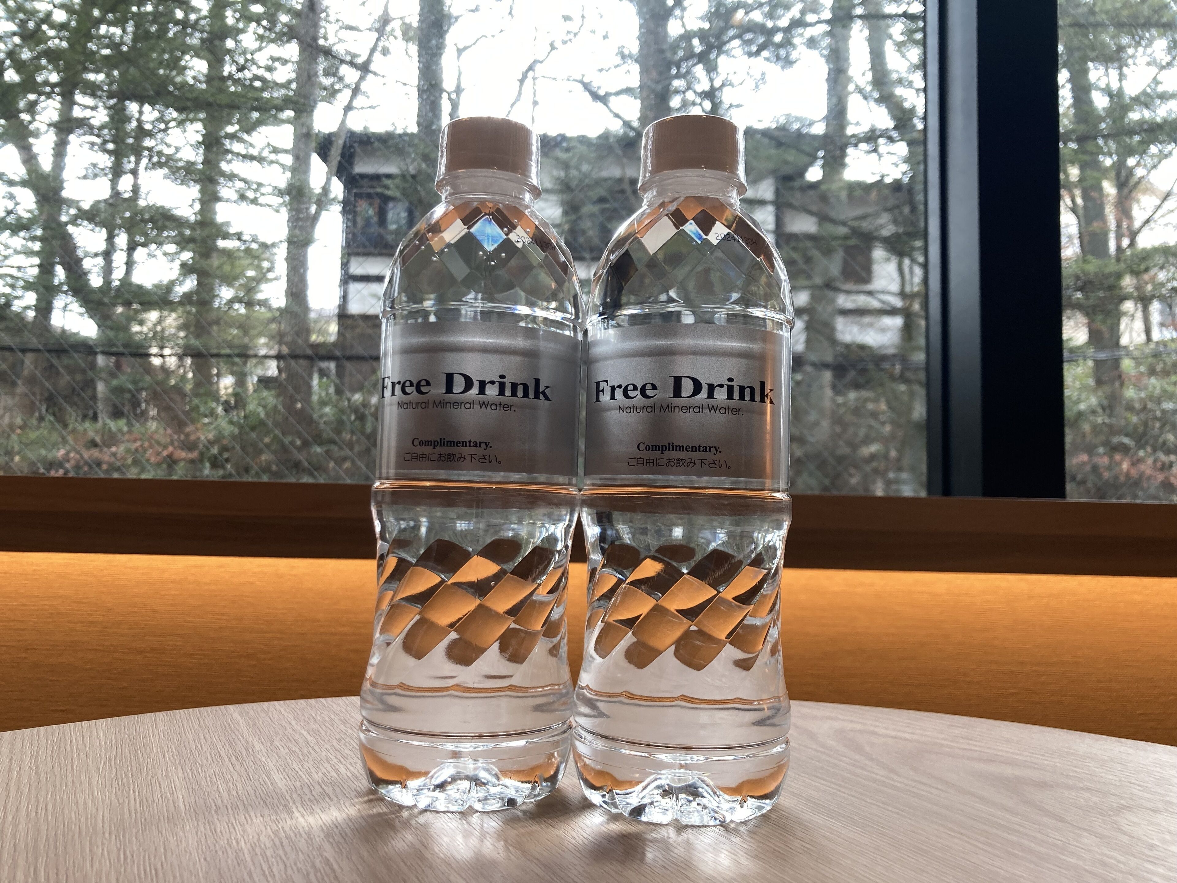 Duas garrafas de água 'Free Drink' com design texturizado, colocadas sobre uma superfície de madeira perto de uma janela com vista para árvores.
