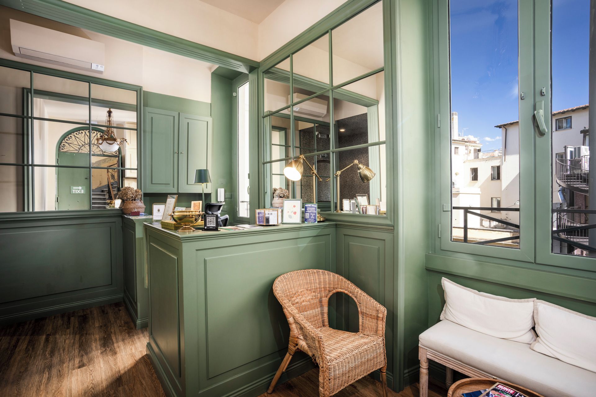 Quarto com paredes em painel verde, uma cadeira de vime, um sofá branco e uma janela com vista para edifícios.