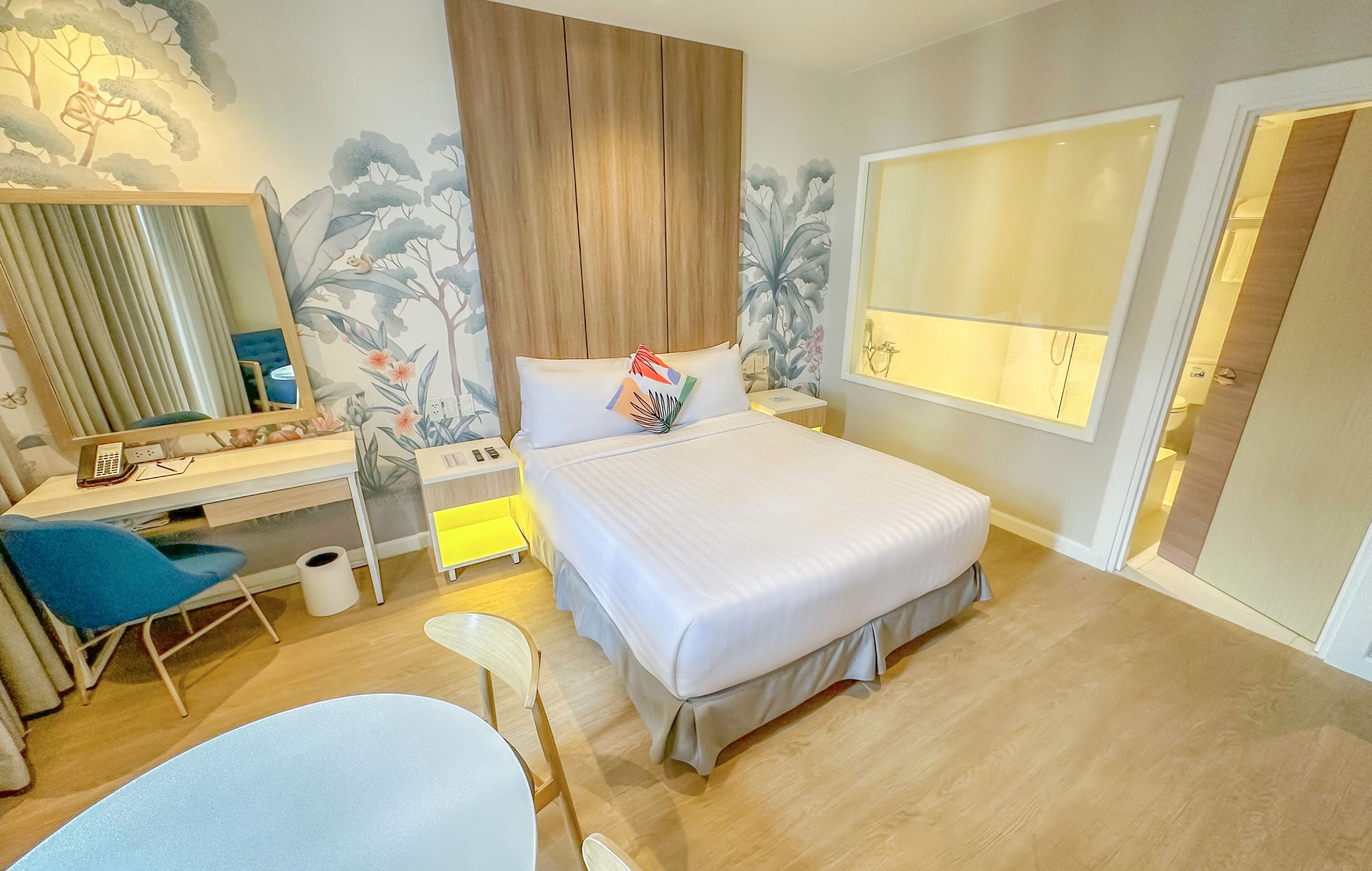 Quarto de hotel com cama, escrivaninha, cadeira e uma janela grande.