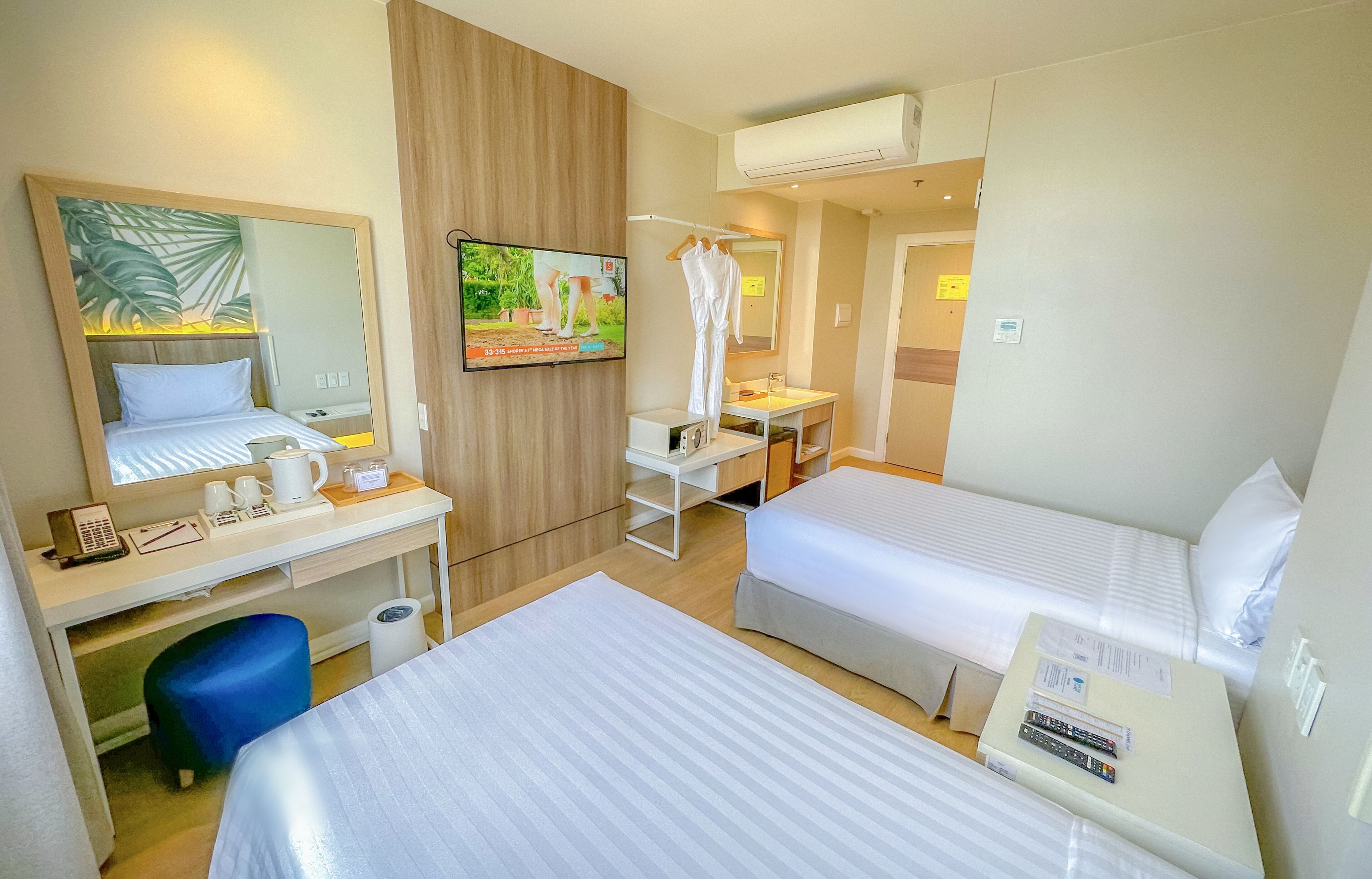 Quarto de hotel com duas camas, uma escrivaninha, uma cadeira, televisão, espelho e ar condicionado.