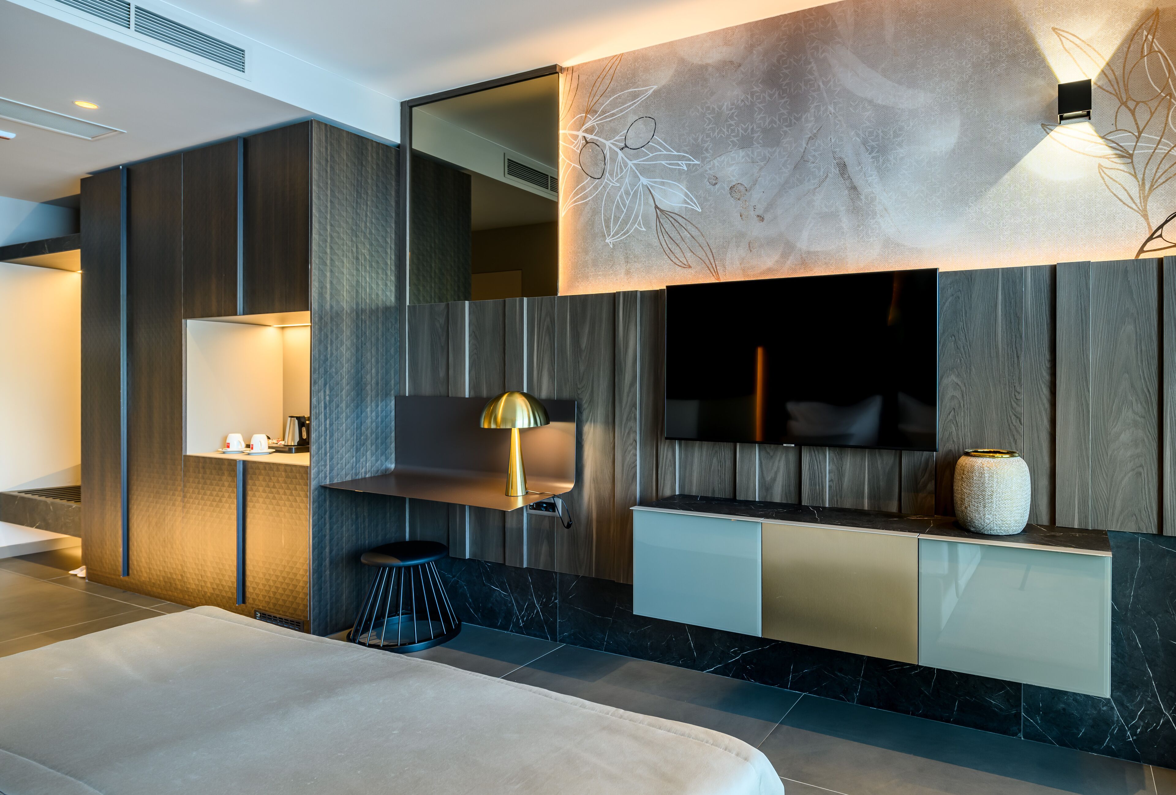 Quarto de hotel moderno com uma cama grande, área de estar com uma luminária, televisão e um detalhe decorativo na parede.