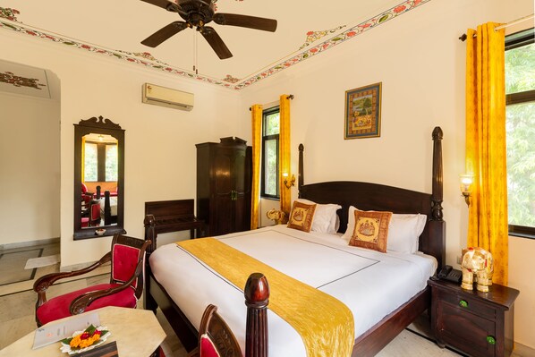 Quarto de hotel com uma cama grande, mesa de cabeceira de madeira, escrivaninha com cadeira, ventoinha no teto e janela com cortinas amarelas.
