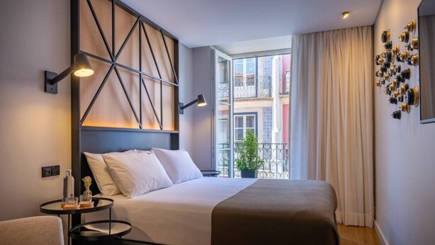 Quarto de hotel moderno com uma cama grande, uma pequena mesa de apoio e vista para a cidade através de uma varanda.