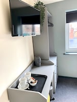 Quarto de hotel com televisão de tela plana na parede, uma mesinha com um suporte contendo uma xícara, uma tigela e alguns petiscos, um chaleira e uma janela com vista para um edifício.