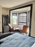 Quarto com cama grande, duas cadeiras, uma mesinha, guarda-roupa e janela com persianas.