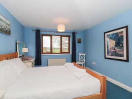 Quarto com uma cama grande, janela com cortinas, mesa de cabeceira com abajur e um quadro emoldurado na parede.