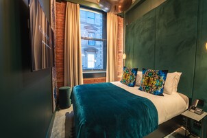 Quarto de hotel moderno com uma cama grande, paredes verdes e vista para um edifício através de uma janela com cortinas.