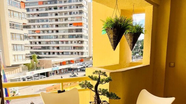 Uma varanda com uma mesa e cadeiras brancas, uma árvore bonsai e vasos suspensos com folhagem.