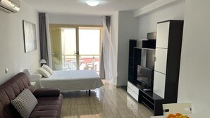 Quarto de hotel moderno com cama, sofá, televisão e área de refeições com frutas.