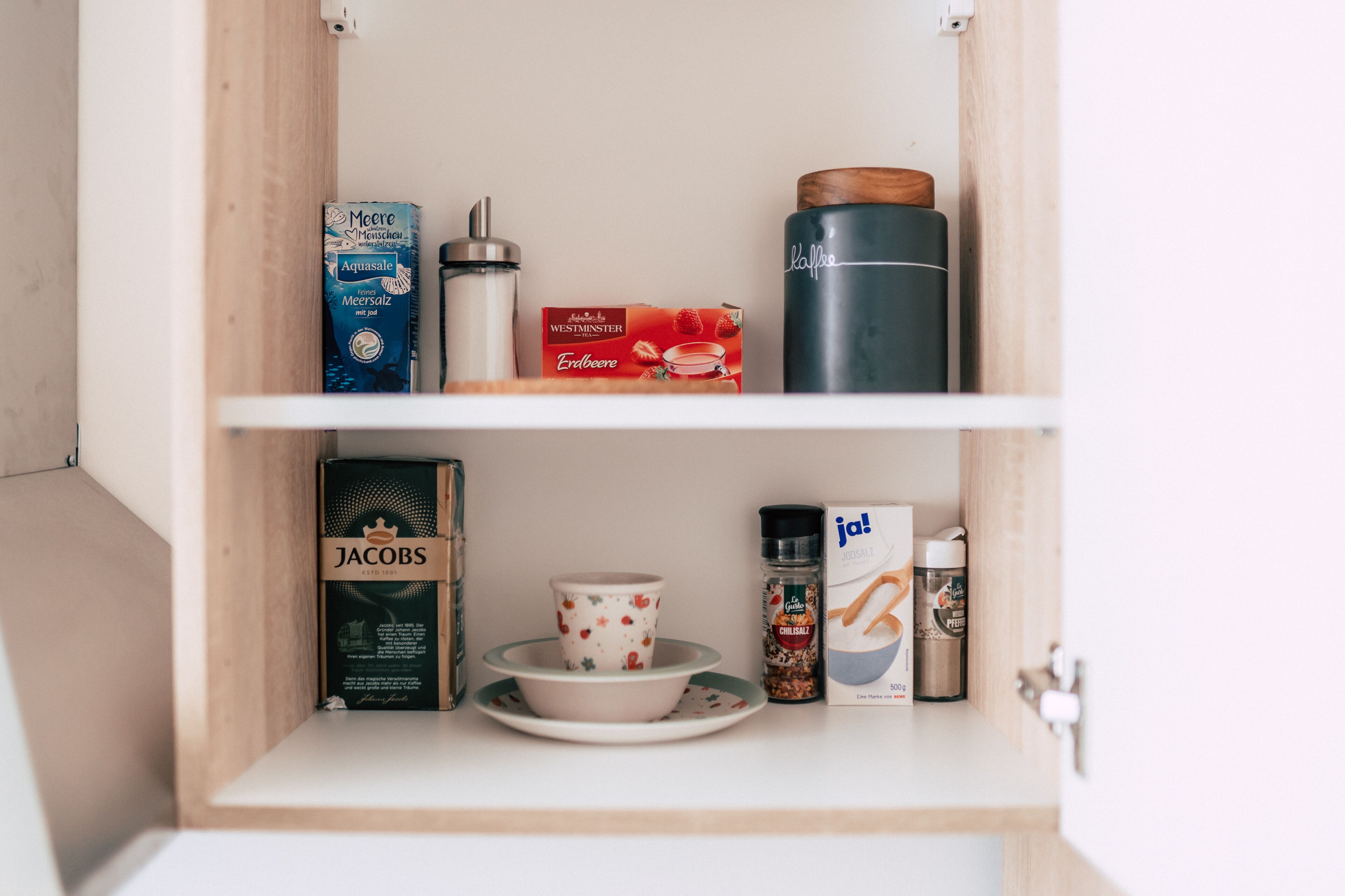 Uma prateleira com uma caixa de café Jacobs, uma xícara e pires, um pote de grãos de café, uma caixa de Westminster Endurose, um recipiente com leite e uma garrafa térmica.