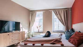 Quarto moderno com televisão de tela plana, cama com várias almofadas, janela com cortinas e cômoda de madeira.