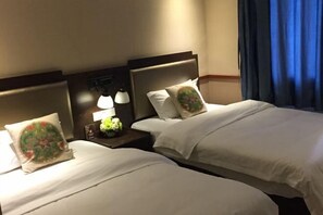 Quarto de hotel com duas camas, cada uma com roupa de cama branca e almofadas decorativas. Há uma mesa de cabeceira com abajur e uma pequena planta.