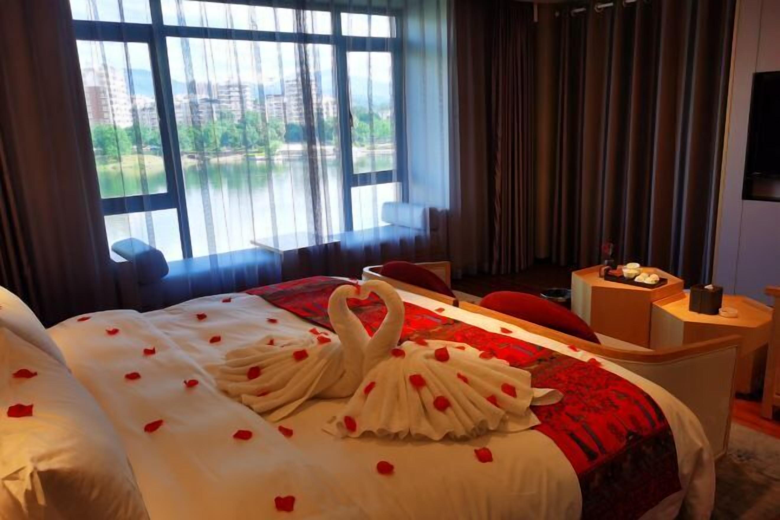 Quarto de hotel com uma cama decorada com pétalas de rosa e toalhas em formato de cisne, vista para o verde e edifícios através de grandes janelas, e uma mesa com luminária e uma pequena mesinha.