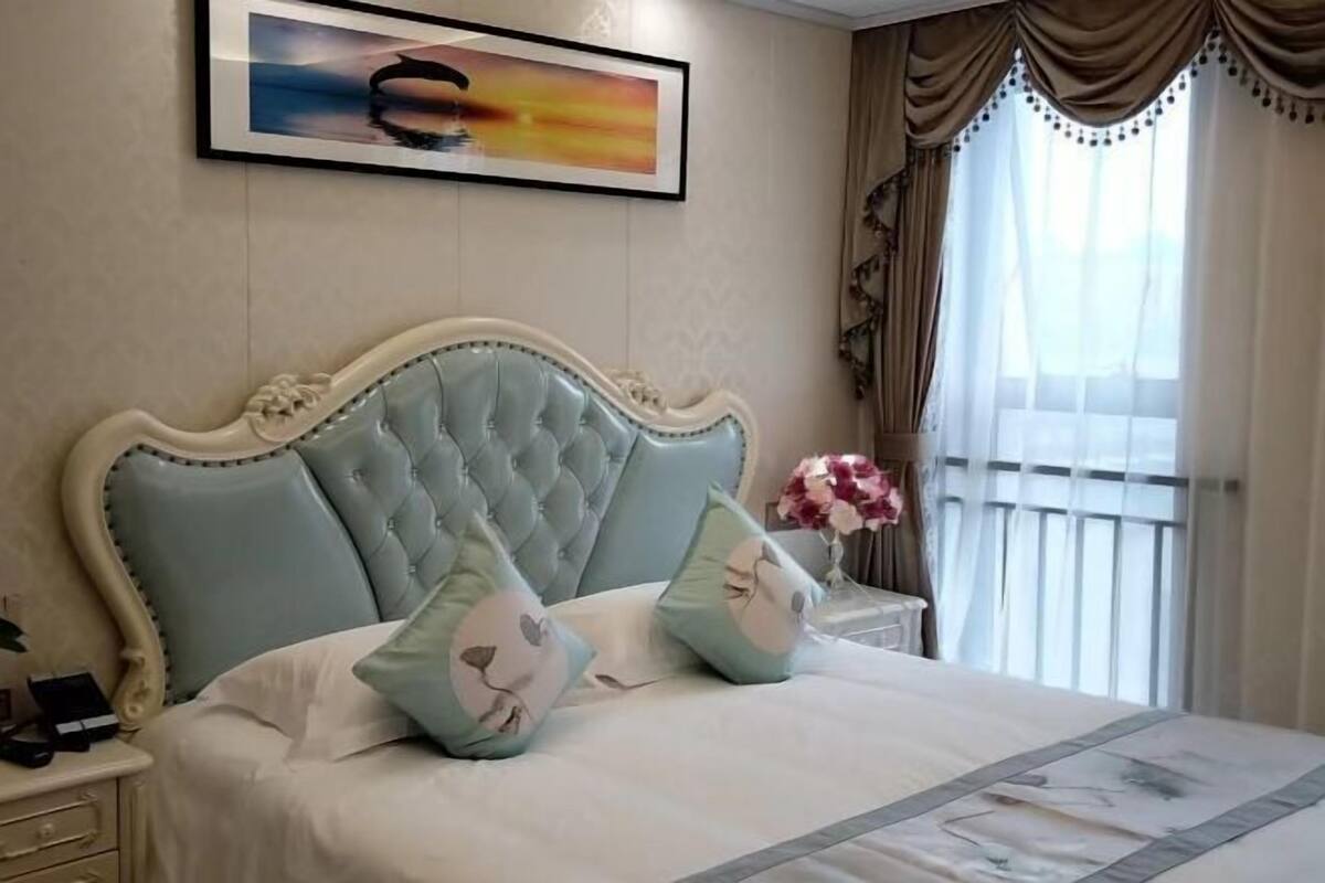 Quarto com cabeceira estofada, cama com almofadas florais, mesa de cabeceira com um vaso e uma moldura na parede.