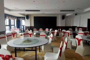 Sala de conferências com mesas redondas e cadeiras brancas com fitas vermelhas, uma tela grande e uma varanda com cortinas.