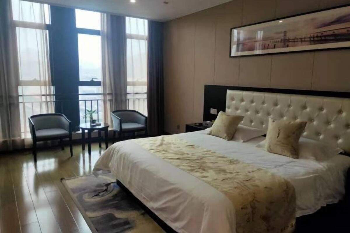 Quarto de hotel com uma cama grande, cabeceira, mesa de cabeceira, uma cadeira, uma mesinha e uma janela com cortinas.